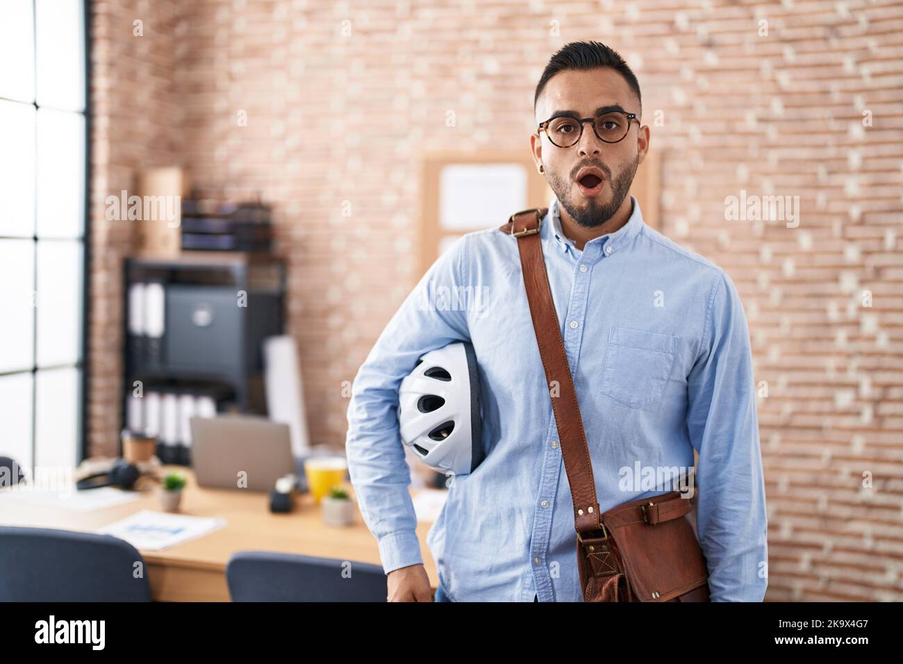 Young hispanic man working at the office holding bike helmet scared and ...