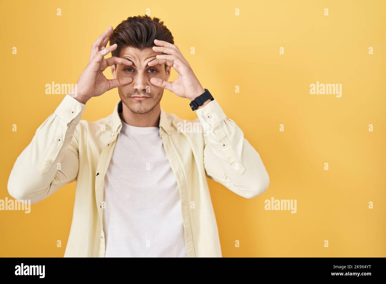 Young hispanic man standing over yellow background trying to open eyes ...