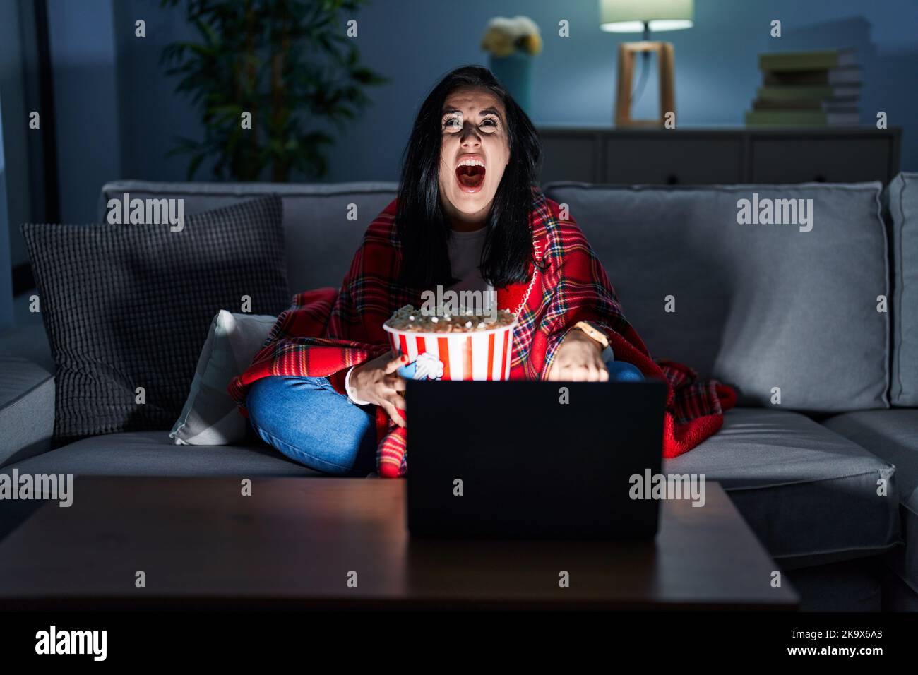Hispanic woman eating popcorn watching a movie on the sofa angry and ...