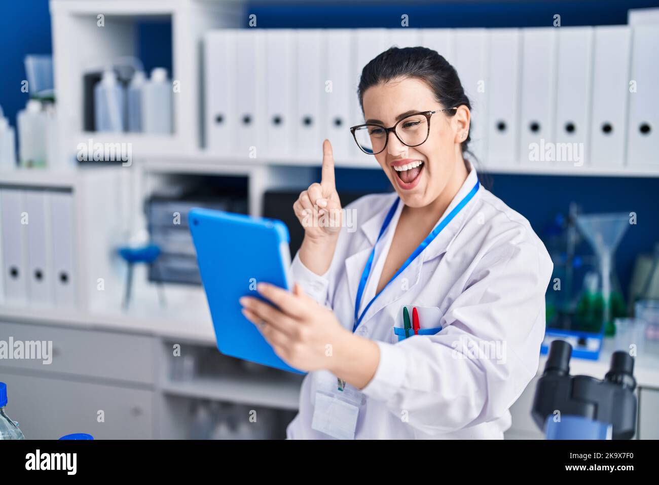 Young brunette woman working at scientist laboratory using tablet smiling with an idea or ...