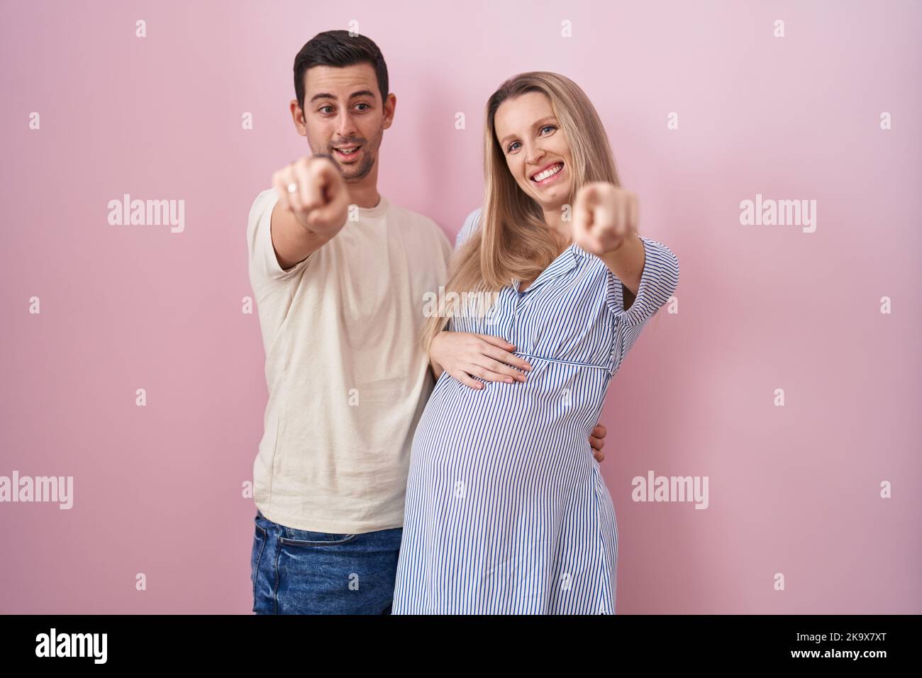 Young couple expecting a baby standing over pink background pointing to ...