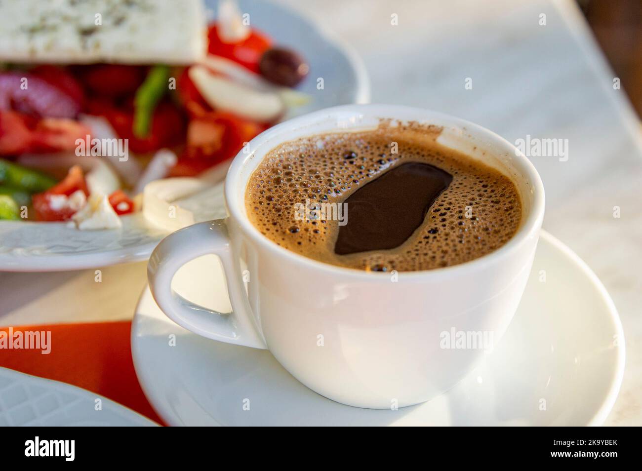 Greek coffee placed on dinning table with colourful Greek salad blurred ...