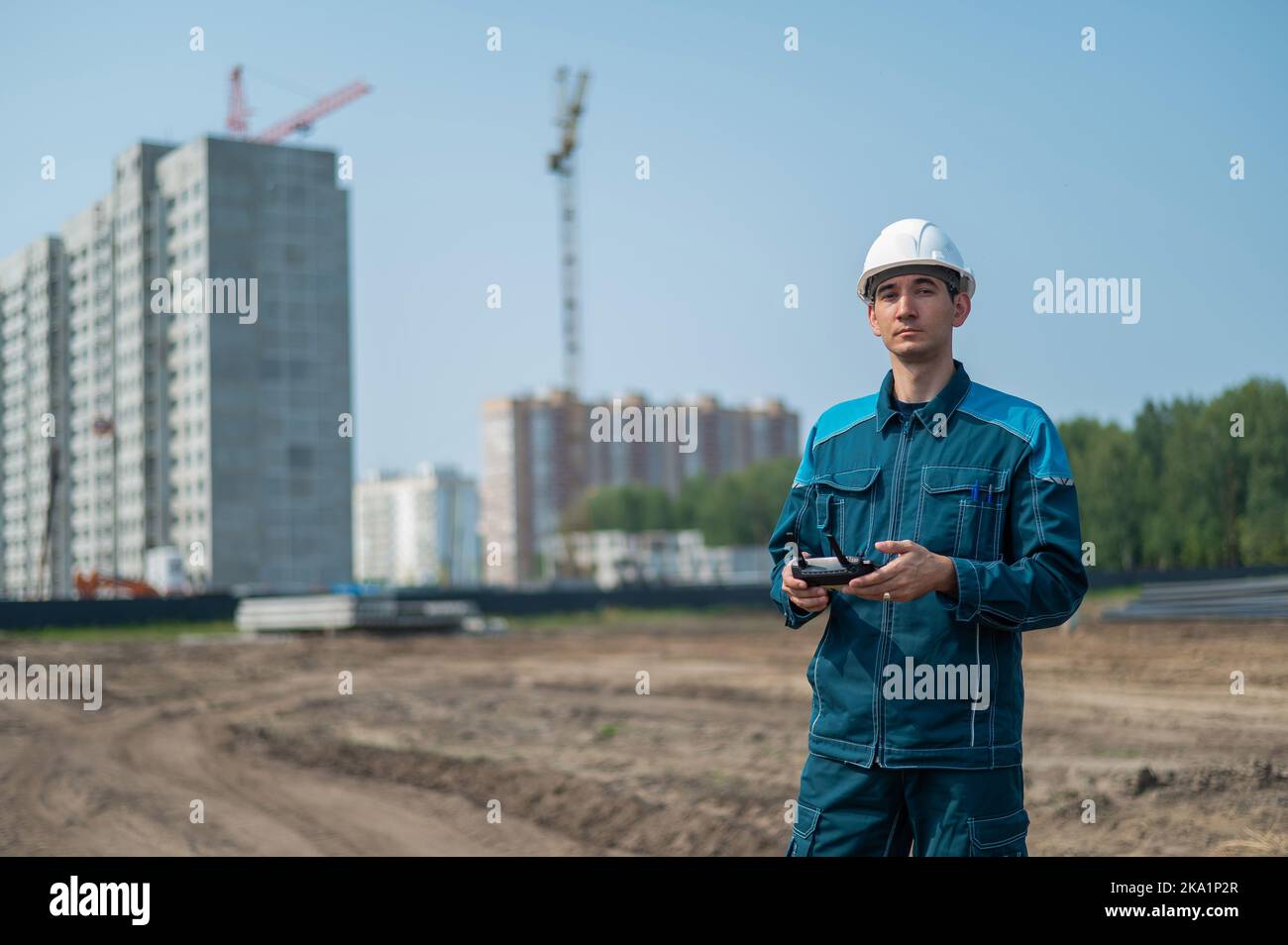 A man in a helmet and overalls controls a drone at a construction site ...