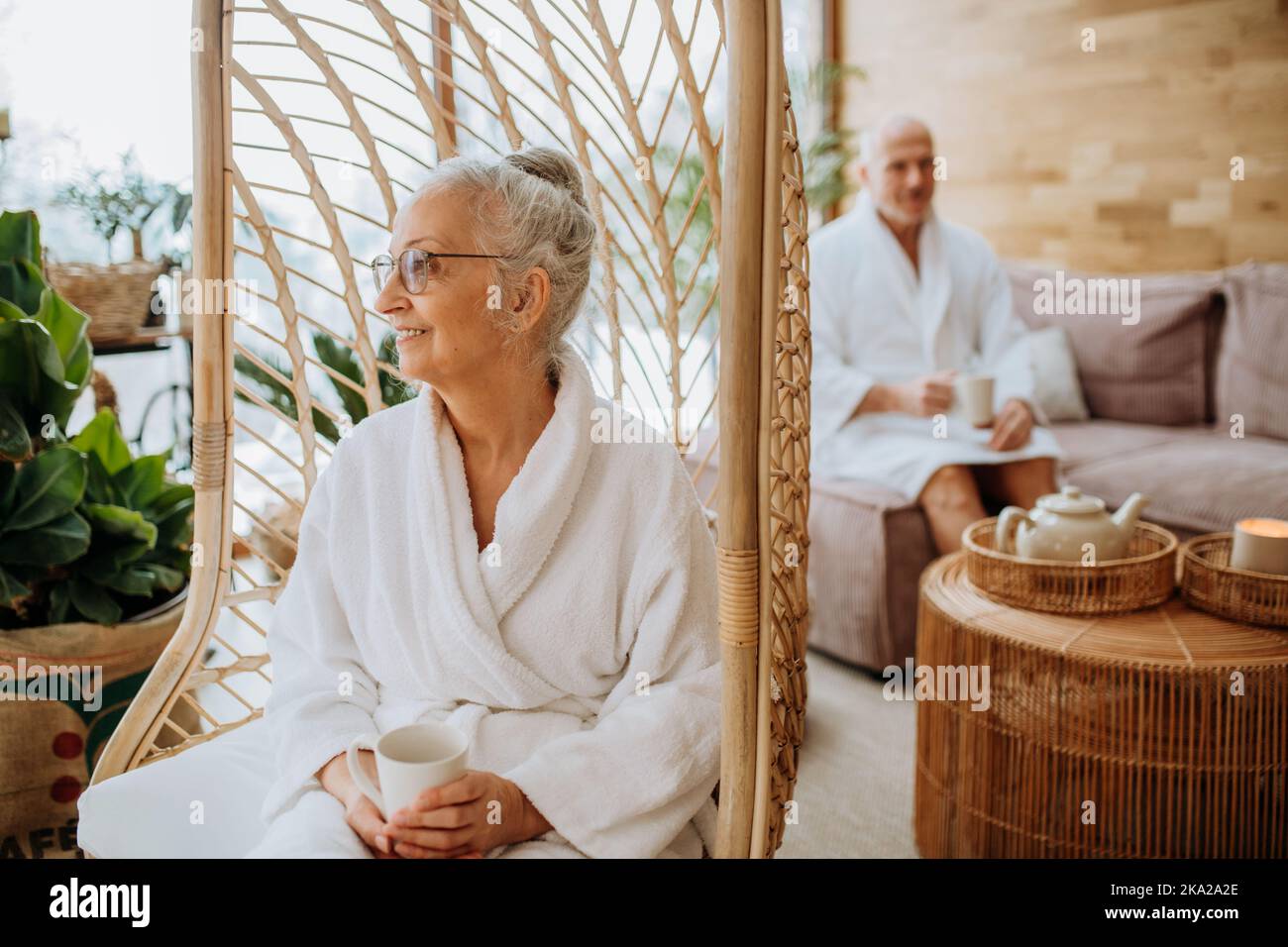 Senior woman sitting on indoor swing with cup of tea, her husband ...