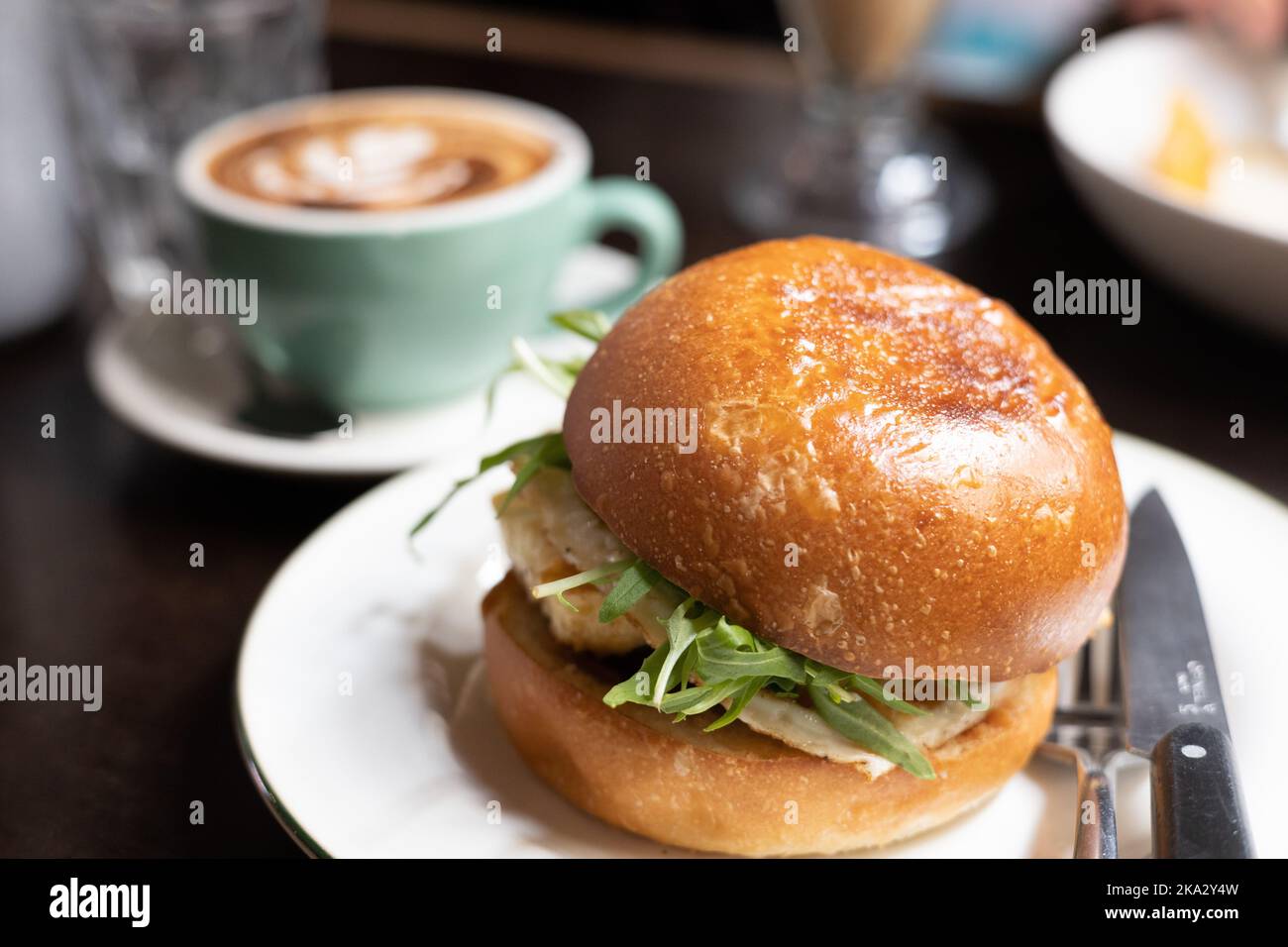 A closeup shot of a hamburger on a white plate with a blurred coffee in ...