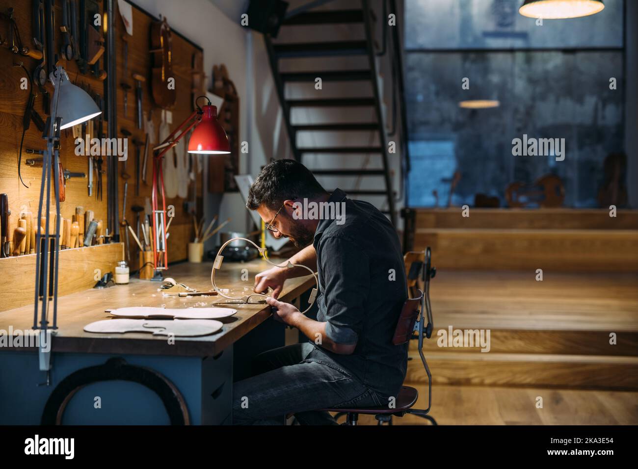 Side view of focused craftsman in glasses sitting at workbench and making new violin starting ...