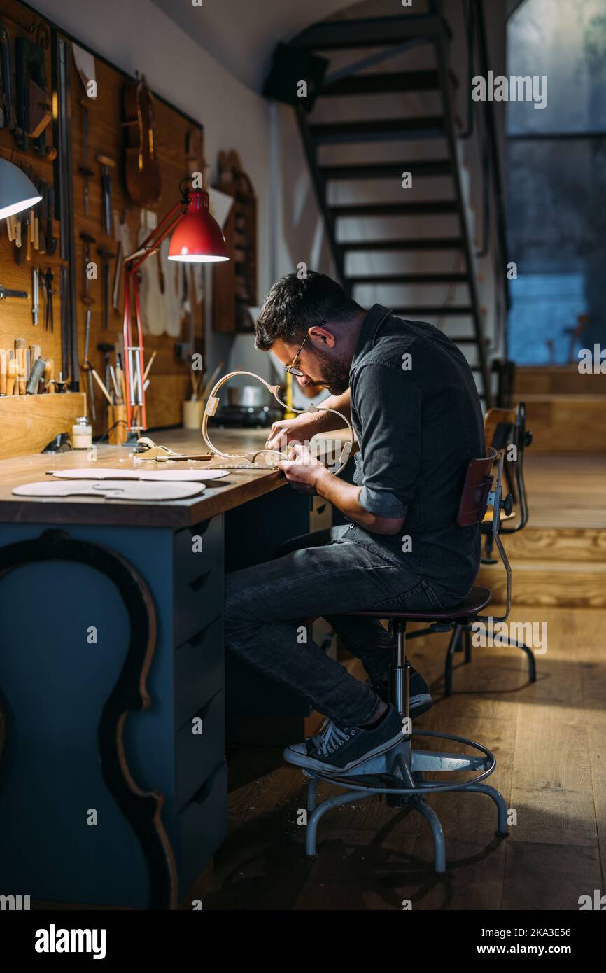 Side view of focused craftsman in glasses sitting at workbench and making new violin starting ...