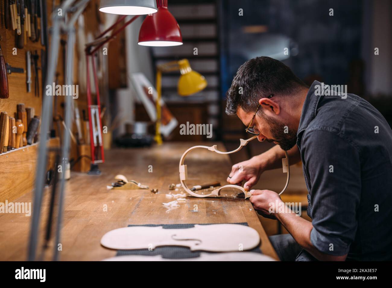 Side view of focused craftsman in glasses sitting at workbench and making new violin starting ...
