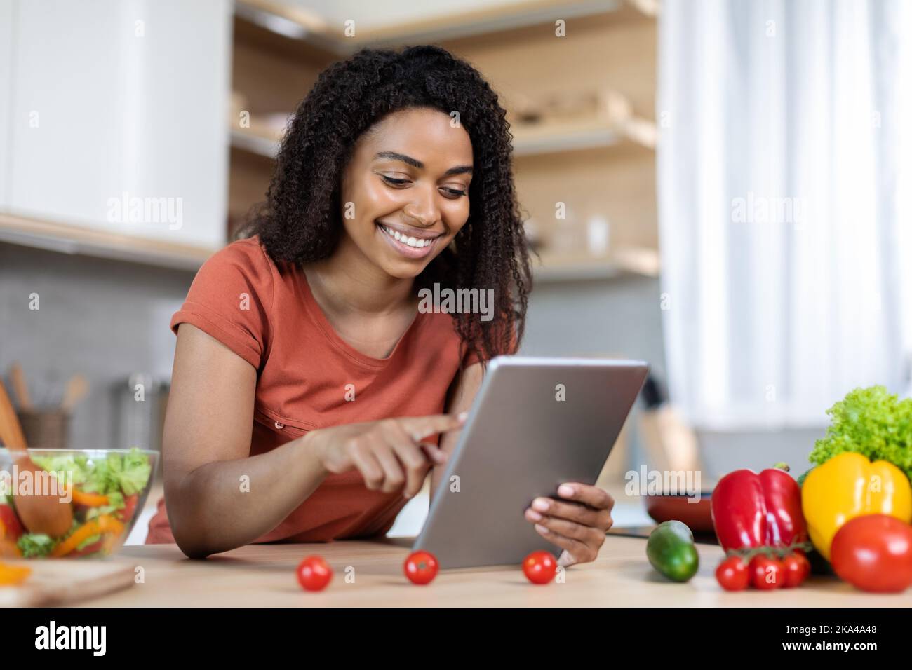 Cheerful young black woman in red t-shirt typing on tablet, read message at table with organic ...