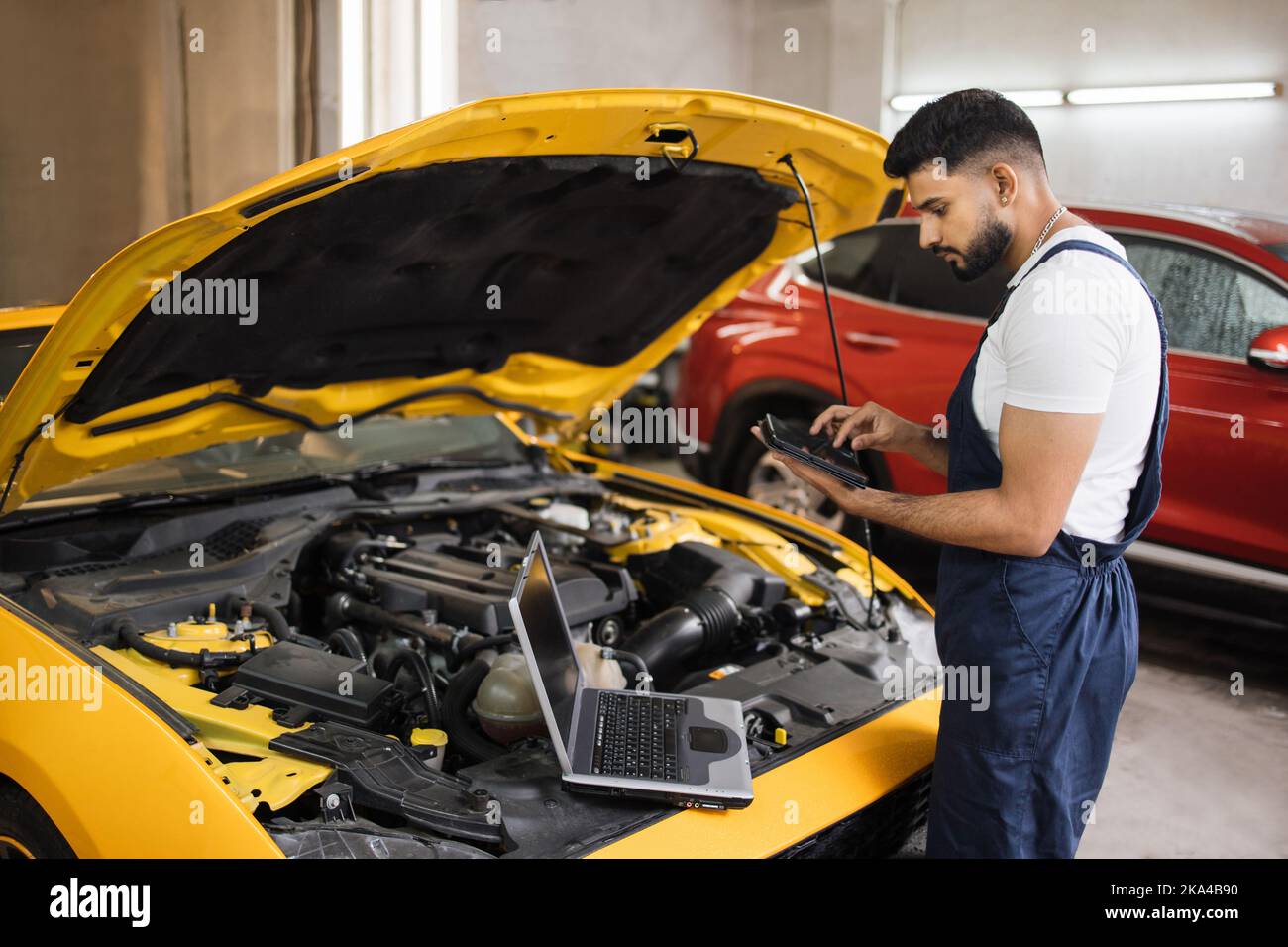 Car service station. Young bearded attractive mechanic man standing and looking under car hood ...
