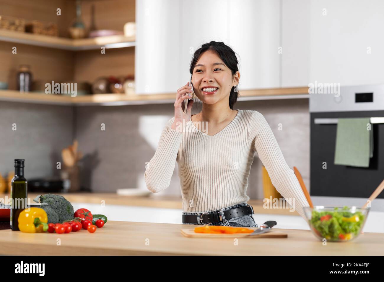 Cheerful pretty asian woman cooking at home, having phone conversation ...