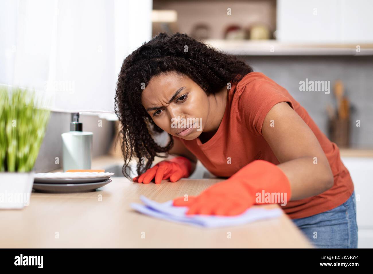 Dissatisfied sad young black lady in red t-shirt, rubber gloves wipes ...