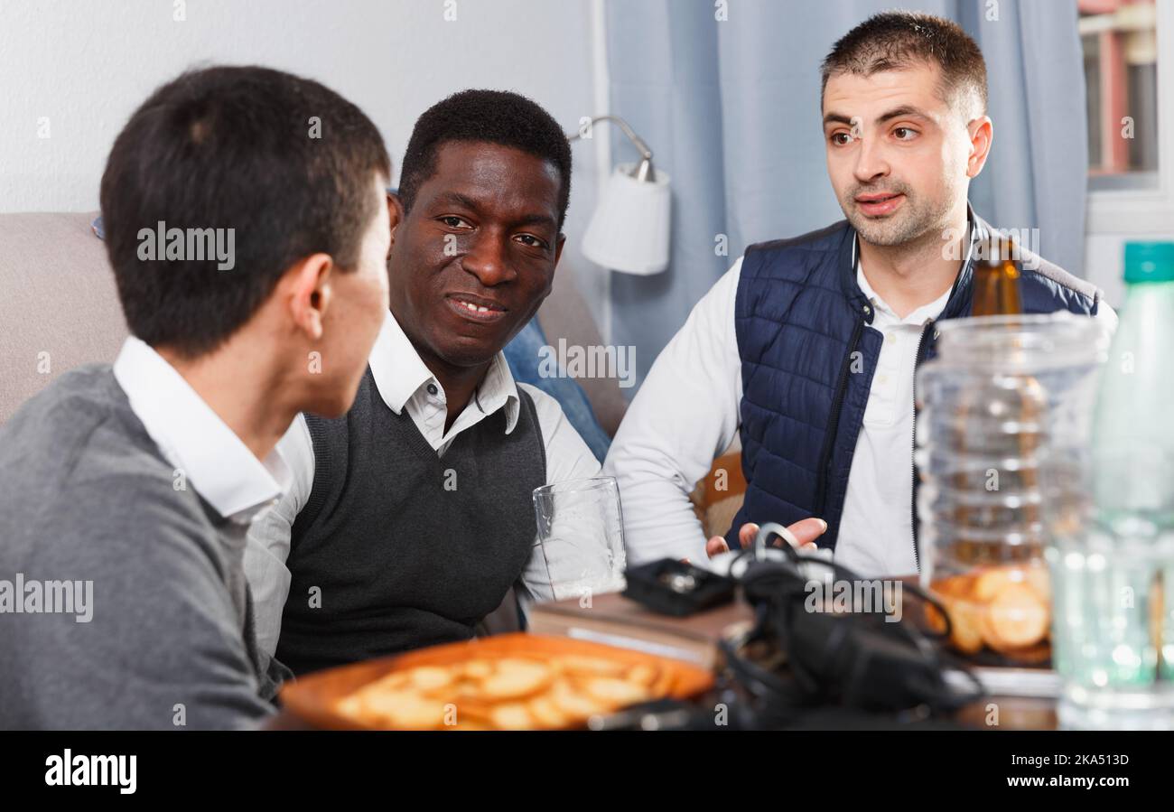 Three cheerful men talking and laughing while drinking beer at home ...