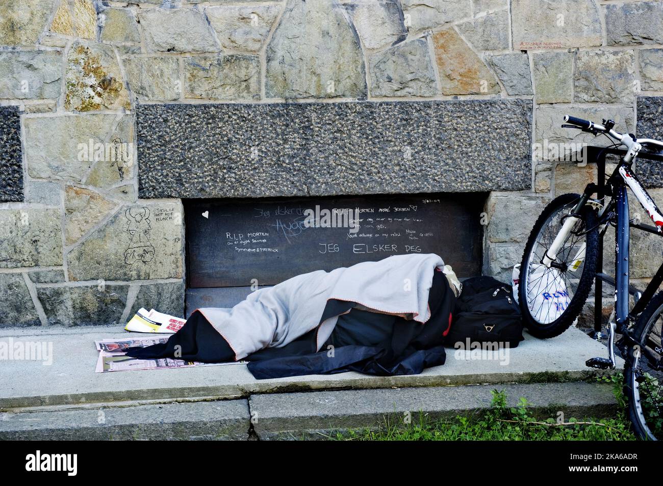 A homeless person sleeps outside with a bicycle parked beside. Writing ...