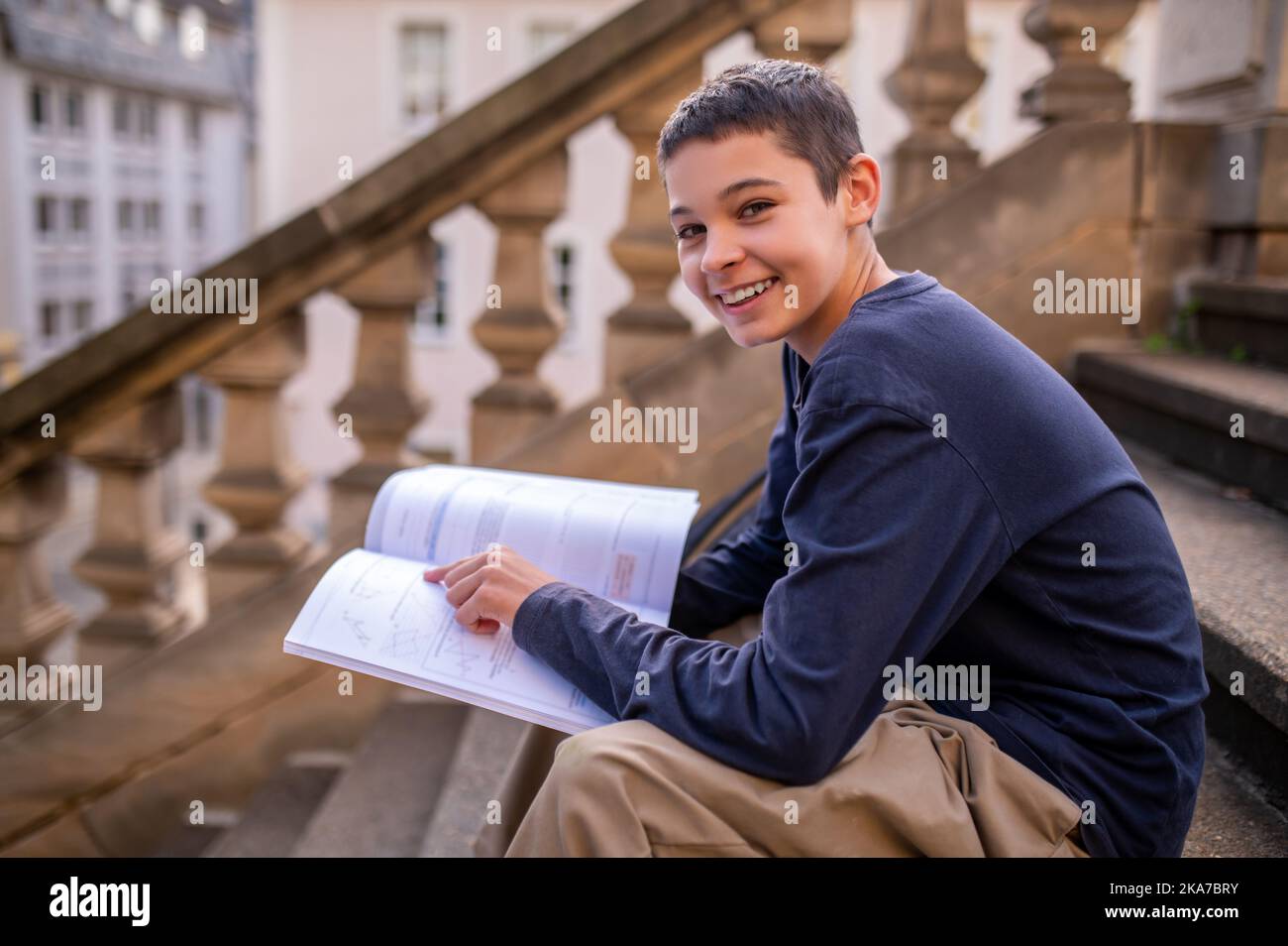 Smiling adolescent doing homework seated on the concrete steps outdoors ...