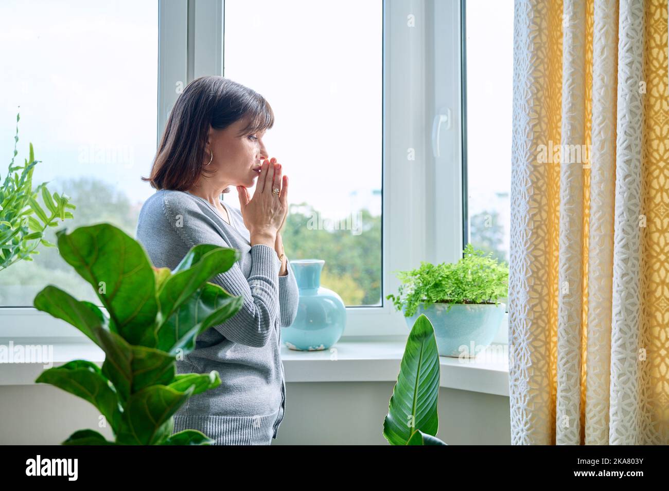 Middle-aged serious woman praying near window at home Stock Photo - Alamy