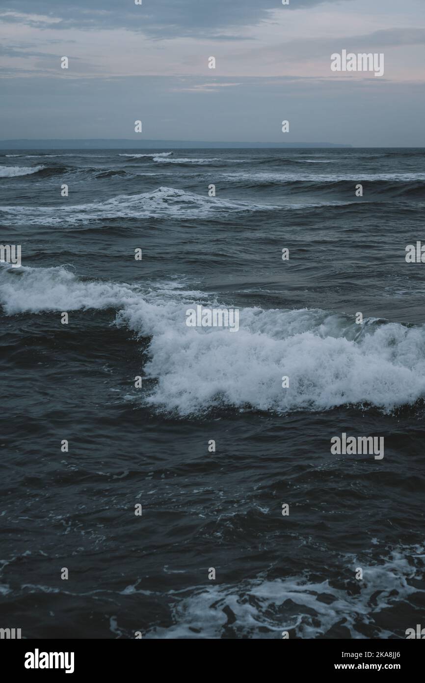 A vertical shot of gray sea waves with foam against a cloudy sunset sky ...