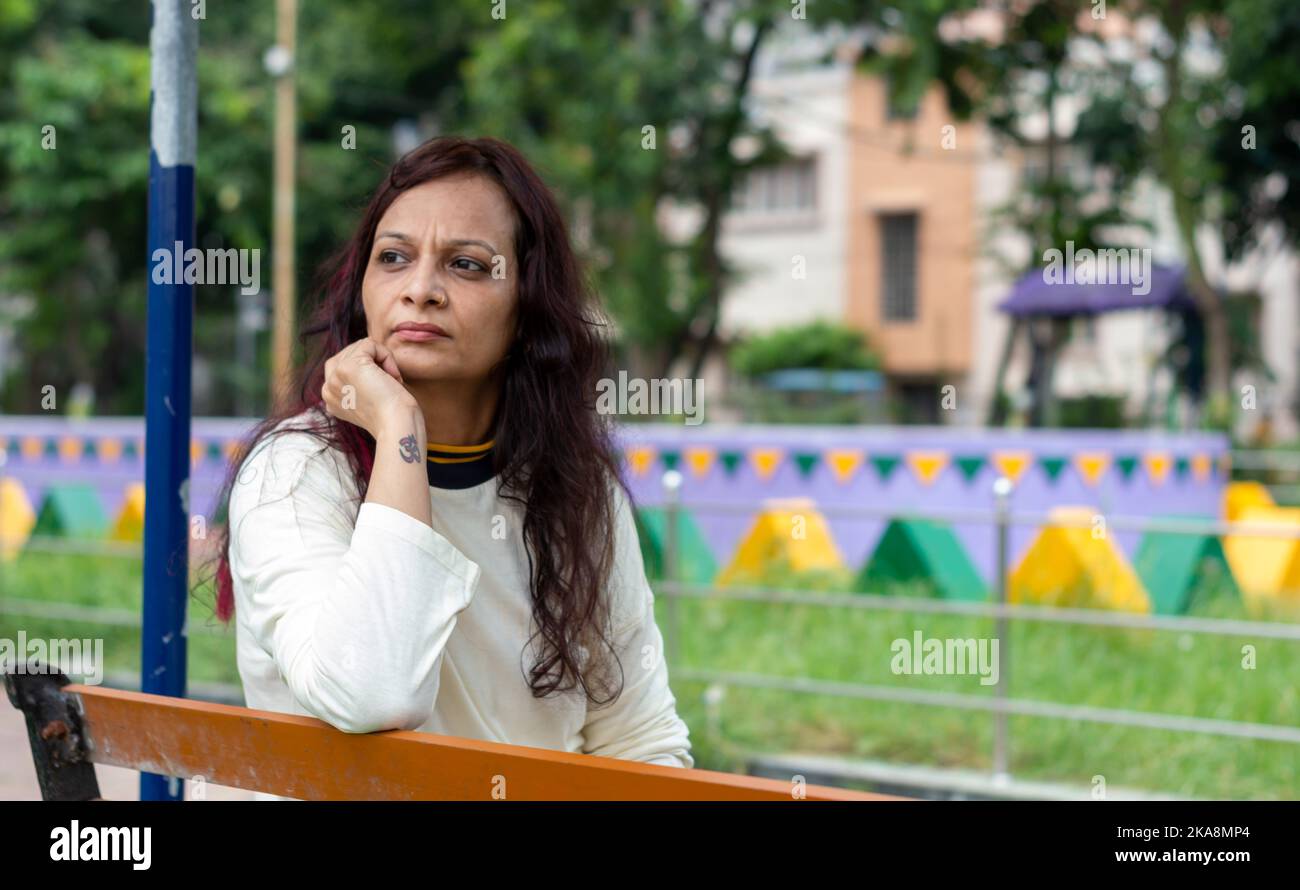 Mid Adult worried Woman sitting on hand on chin pose in a park bench ...