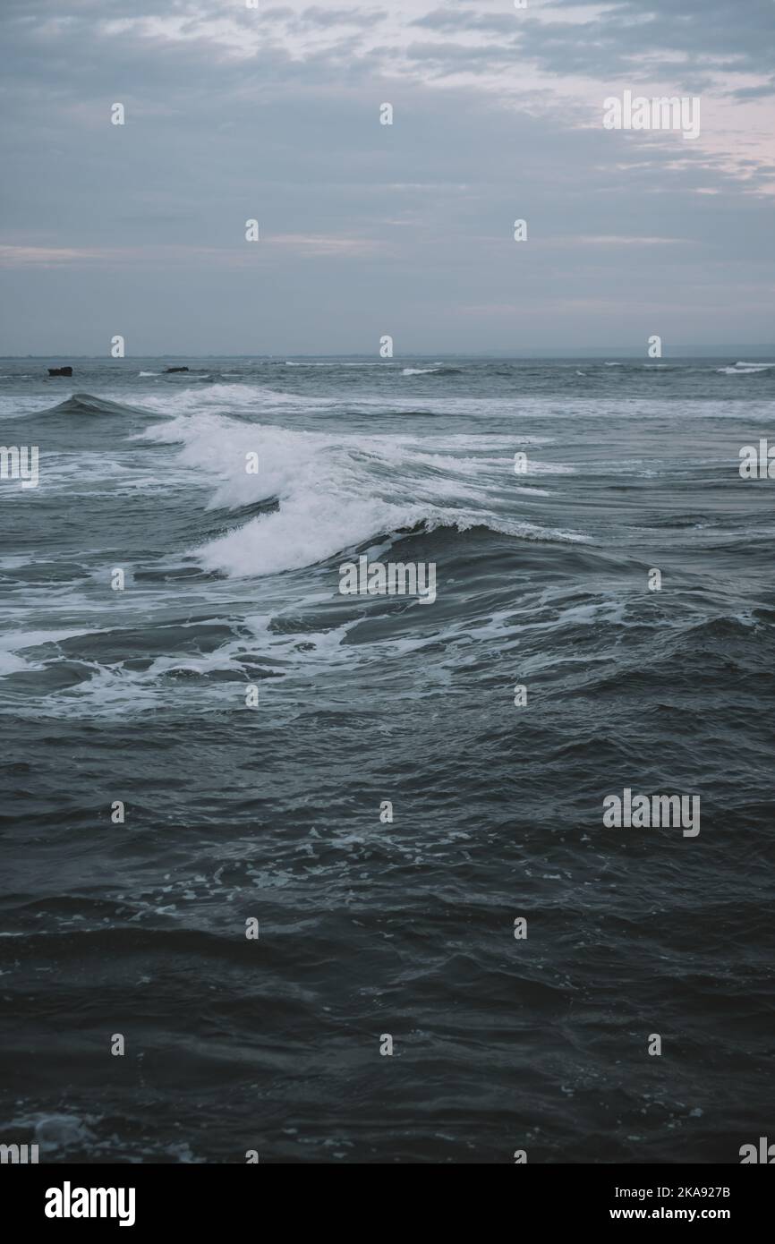 A vertical shot of gray sea waves with foam against a cloudy sunset sky ...