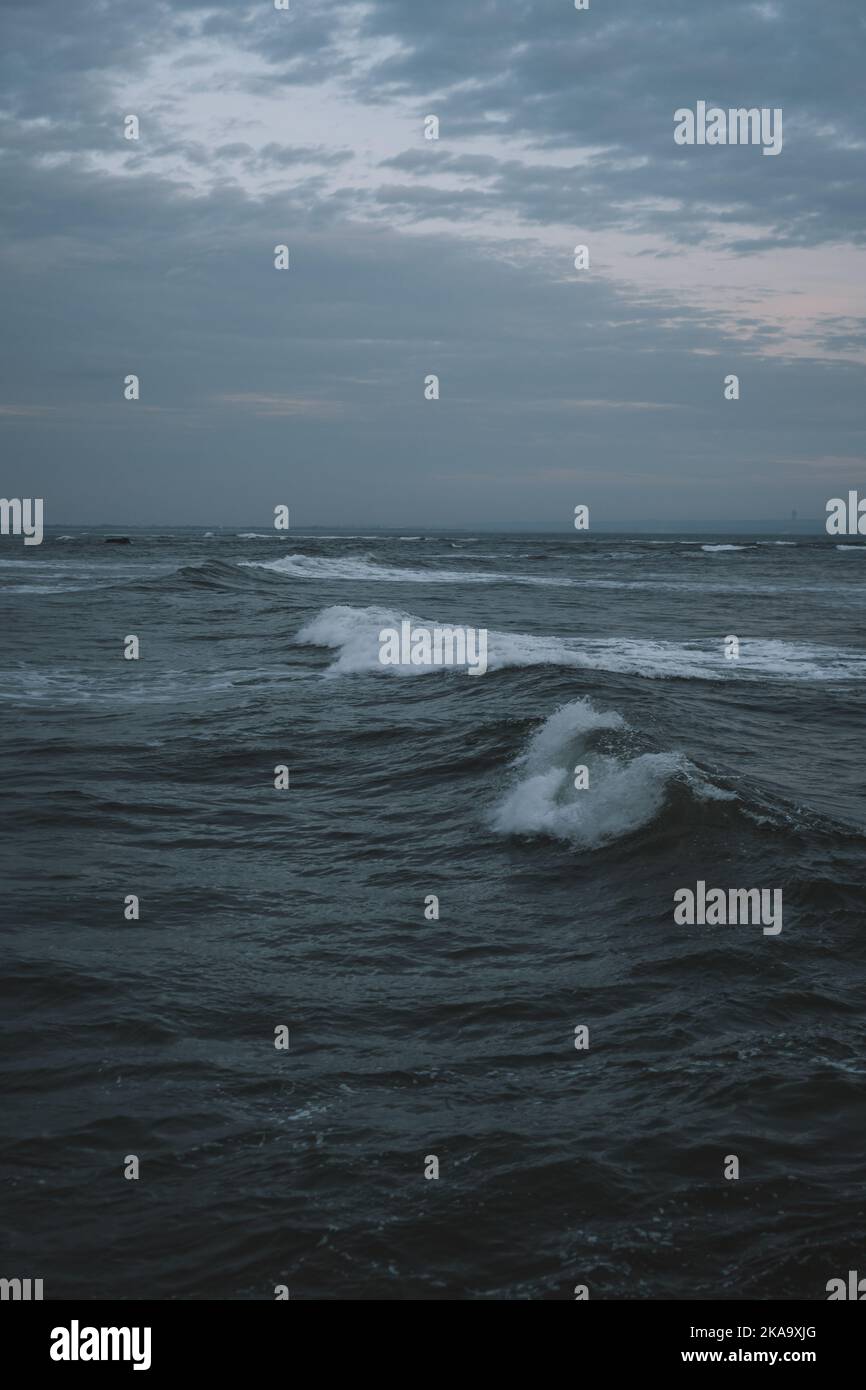 A vertical shot of gray sea waves with foam against a cloudy sunset sky ...