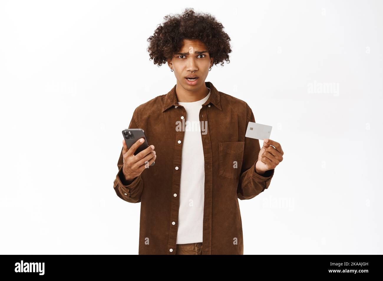 Confused guy holds mobile phone and credit card, looks worried, stands ...