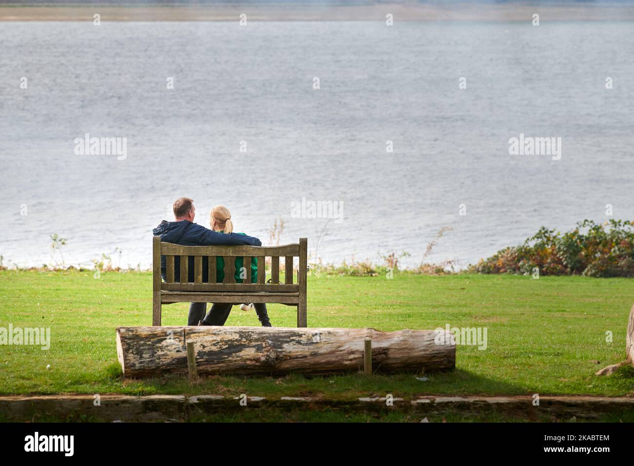 A couple sit close together on a bench beside a lake in an english ...