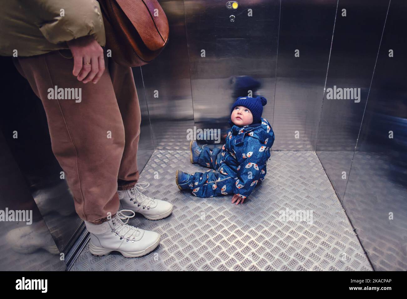 Toddler baby sits on the floor in the elevator next to his mother. The ...