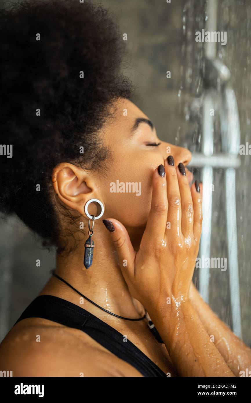 Side view of african american woman washing face under water in shower ...