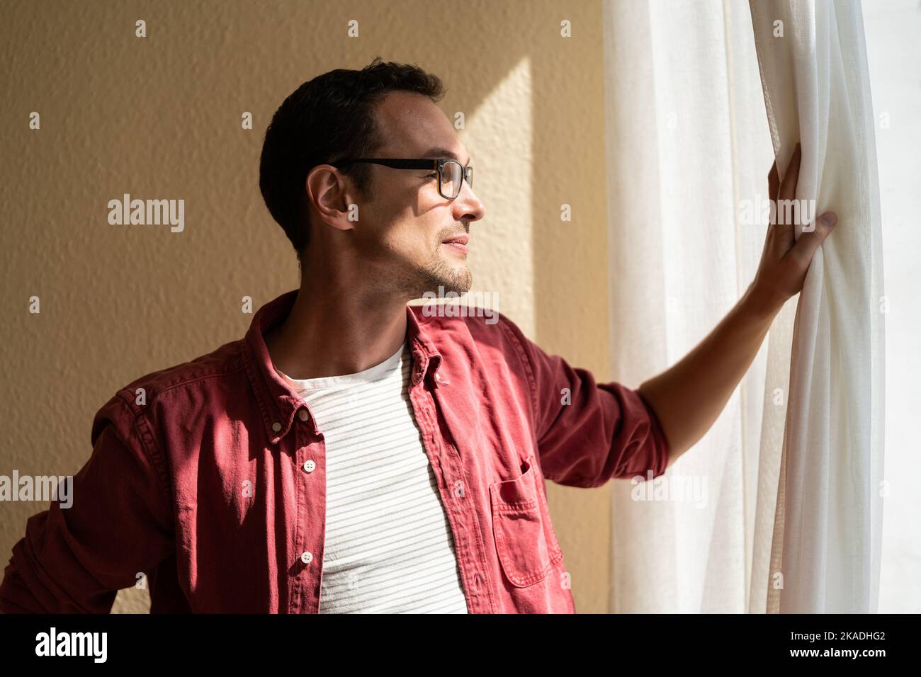 Meeting new day. Young pensive man opening curtains, looking through ...