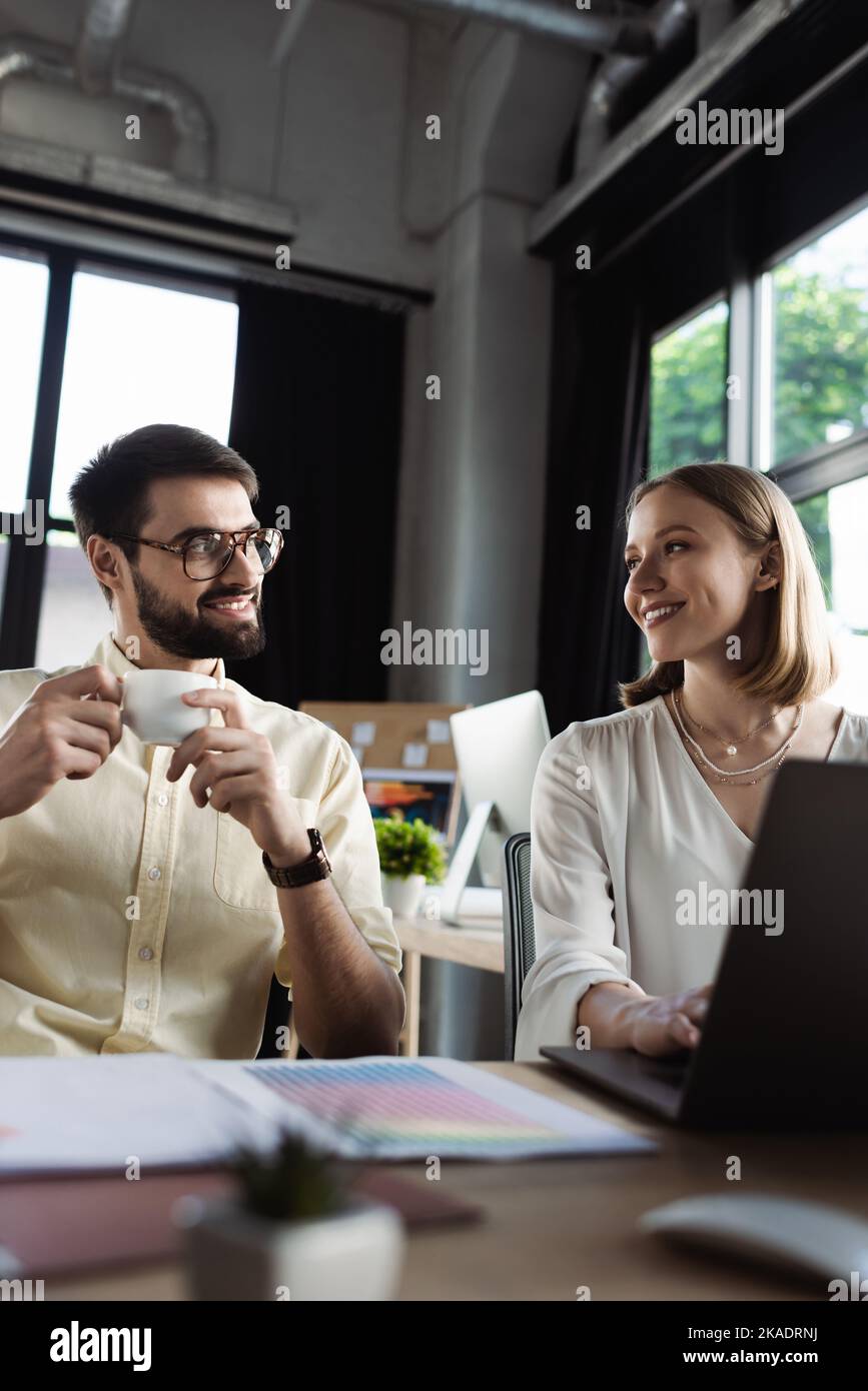 Cheerful intern looking at businessman with cup of coffee near laptop ...