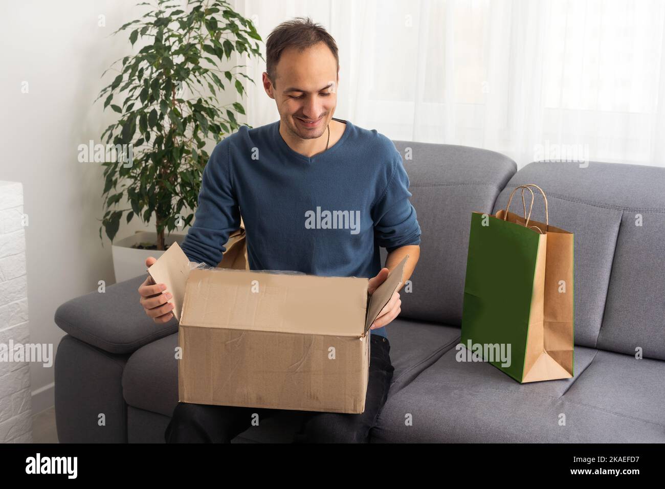 Handsome young man opening box with parcel while sitting on sofa at ...