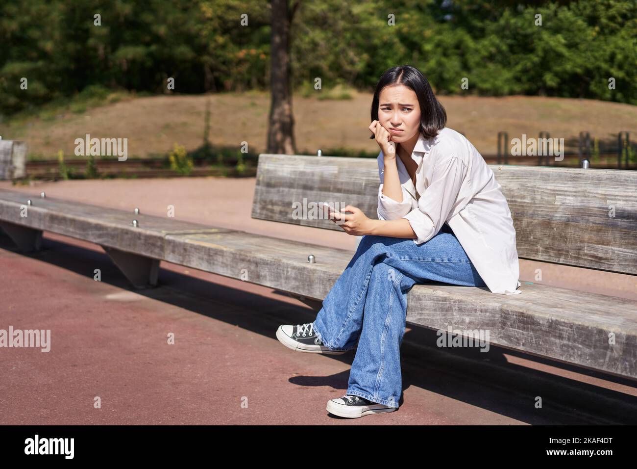 Depressed asian girl sits on bench in park with smartphone, feeling ...