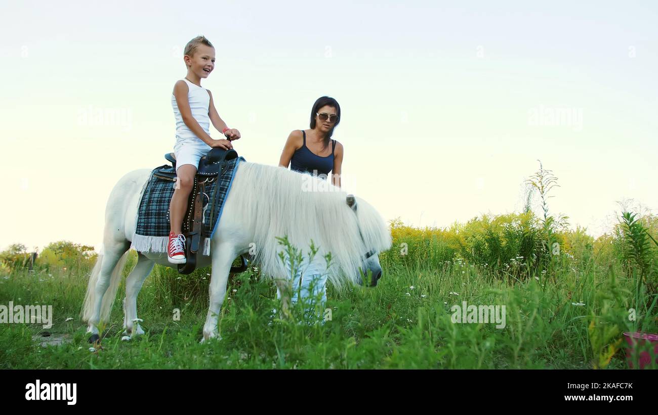A woman and a boy are walking around the field, son is riding a pony ...