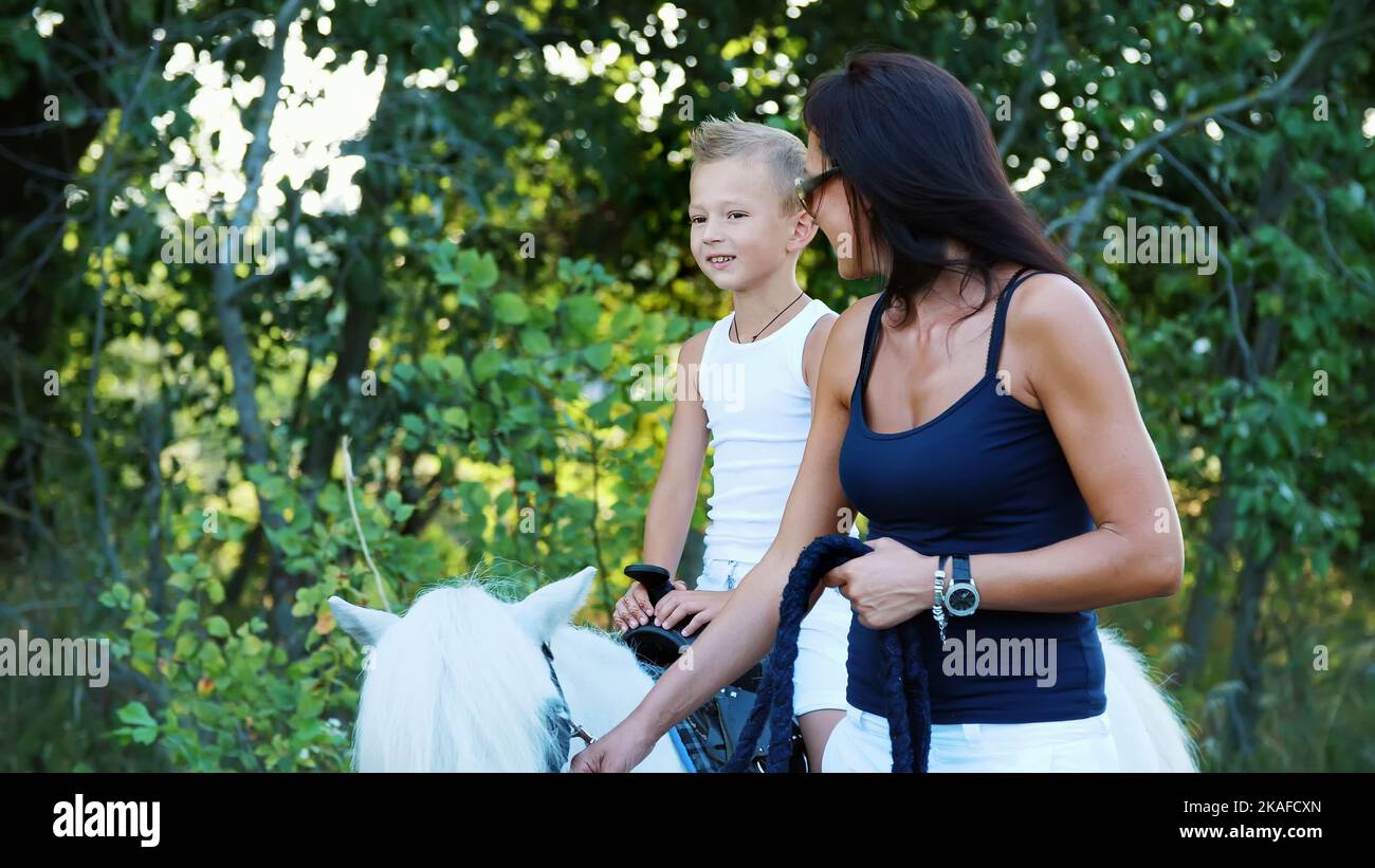 A woman and a boy are walking around the field, son is riding a pony ...