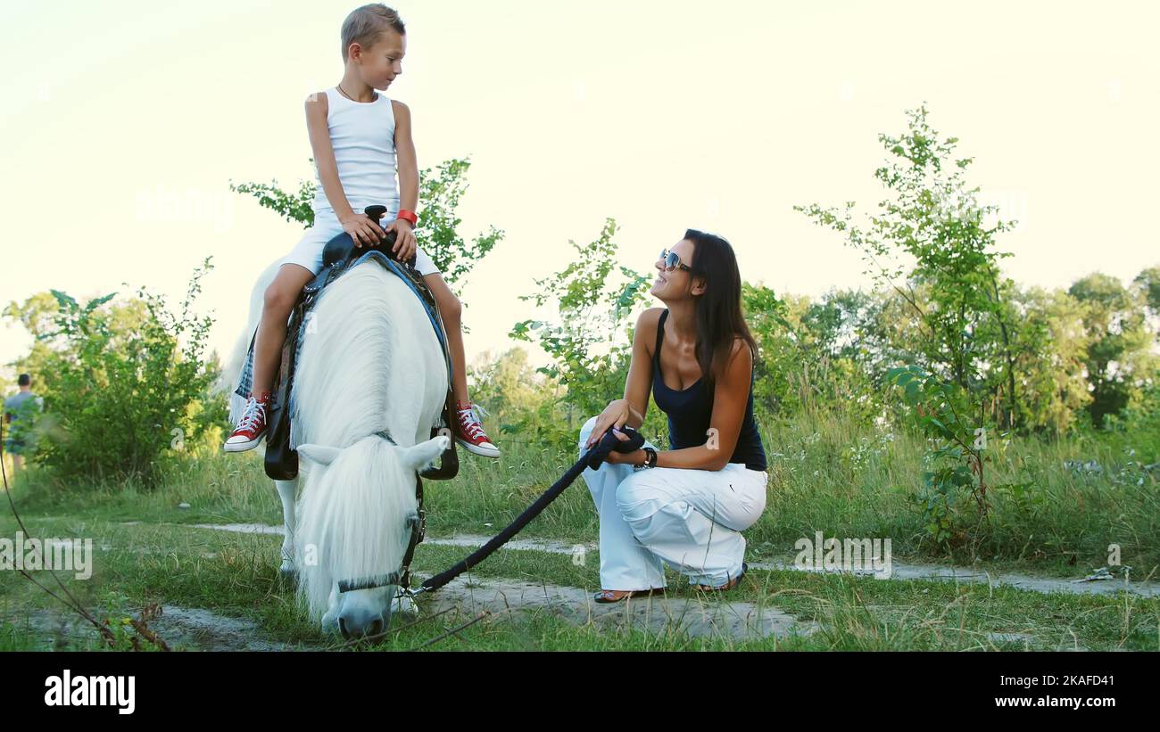 A woman and a boy are walking around the field, son is riding a pony ...