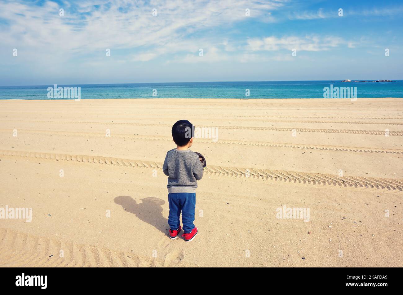 A little boy standing on a sandy shore of the blue sea looking into the ...