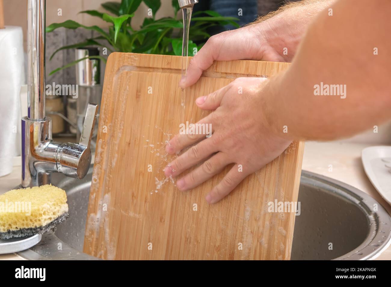 A man washes a wooden bamboo cutting board in the kitchen sink under ...