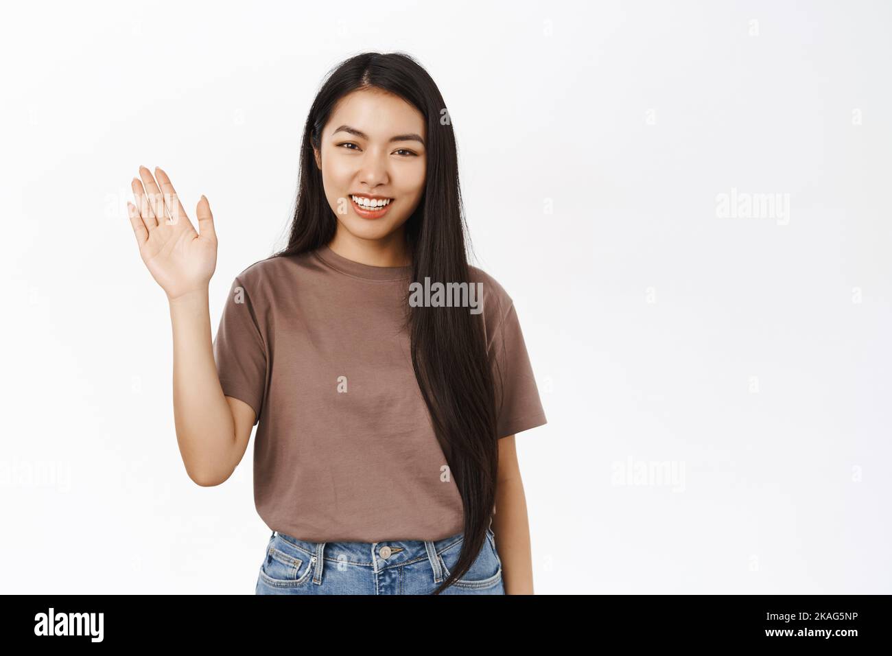 Friendly korean girl waves hand, say hello, hi and smiles at you ...