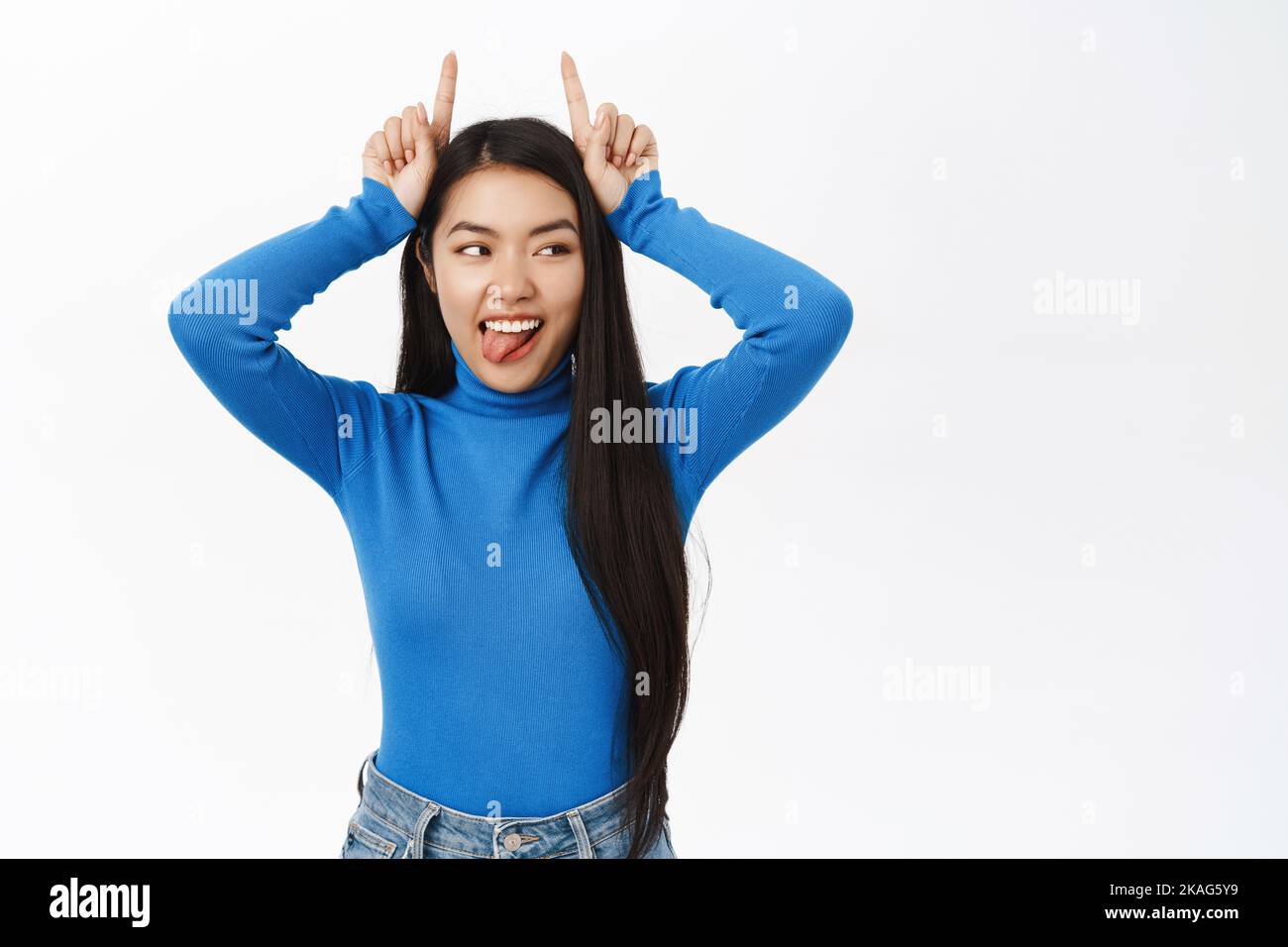 Enthusiastic asian woman shows devil horns, fingers on head, smiles ...