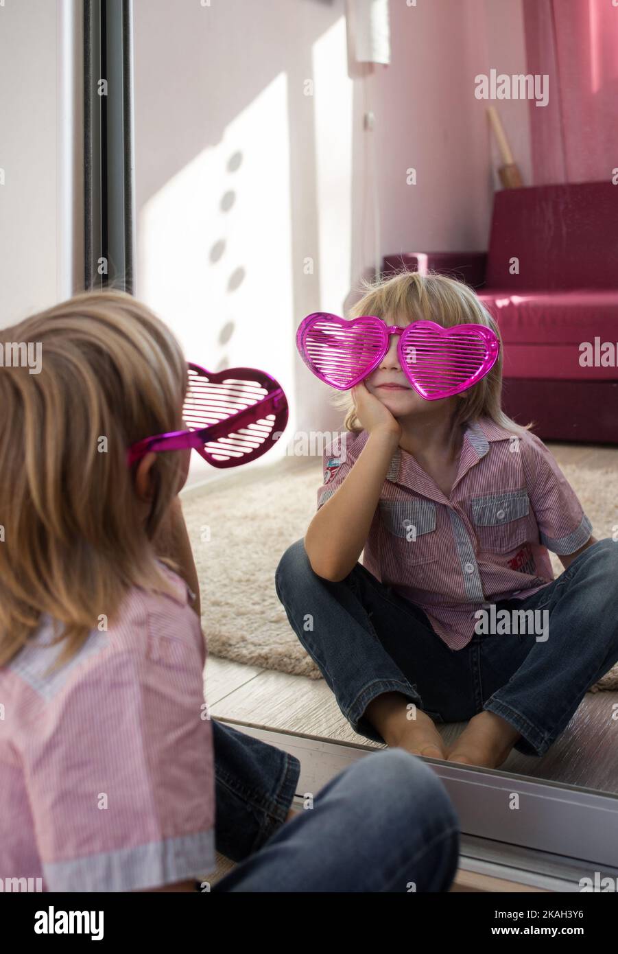 cute stylish boy in huge pink heart-shaped funny glasses sits in a room ...