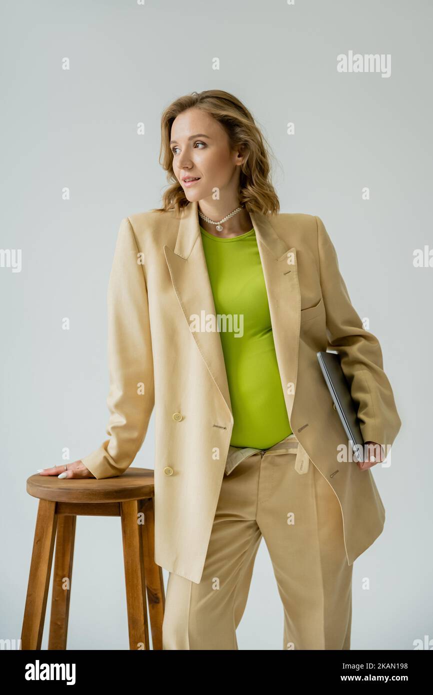 Pregnant woman in suit holding laptop near wooden chair isolated on ...
