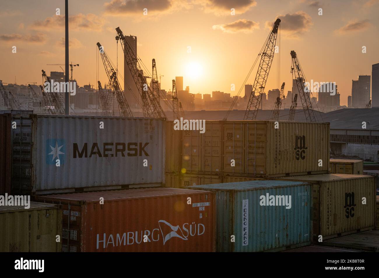 The sun sets behind the urban skyline as port cranes and shipping ...