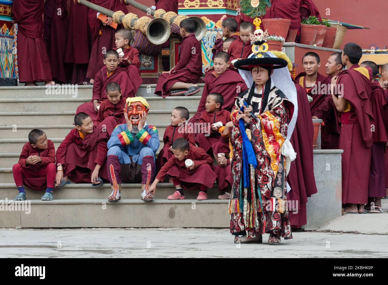 Young tibetan monks sit around a monk wearing achara (joker) mask ...