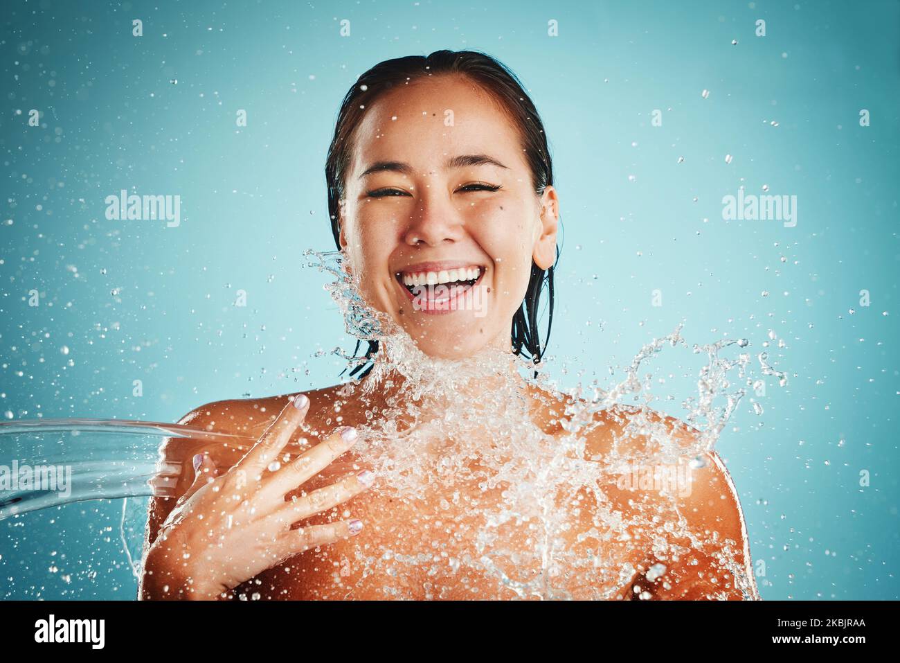 Happy, water splash and woman in studio for skincare, cleaning and beauty on a blue background ...