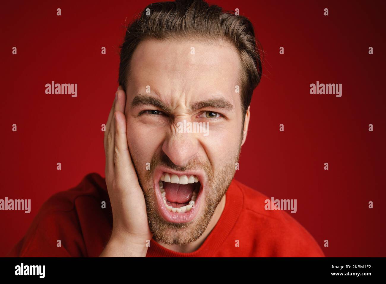 Young bristle man wearing sweater screaming at camera isolated over red ...