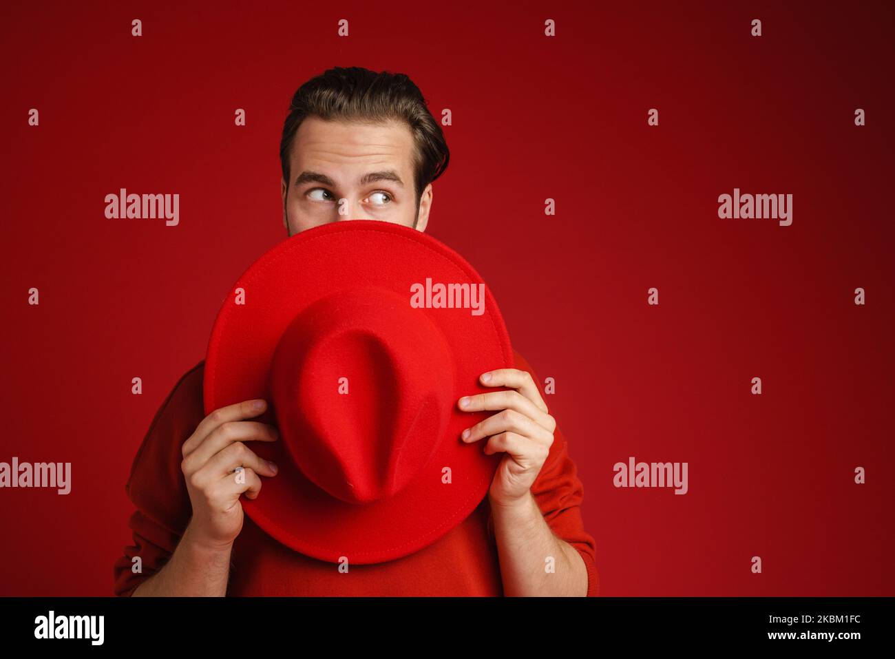 Young white man looking aside while posing with hut isolated over red ...