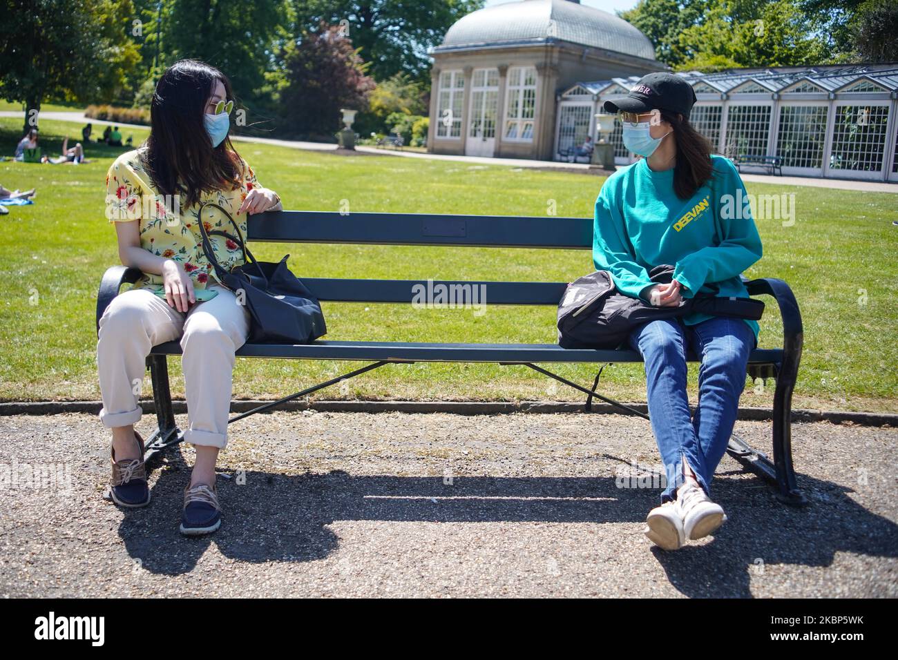 Two friends wearing face masks sit on a bench as they maintain social ...