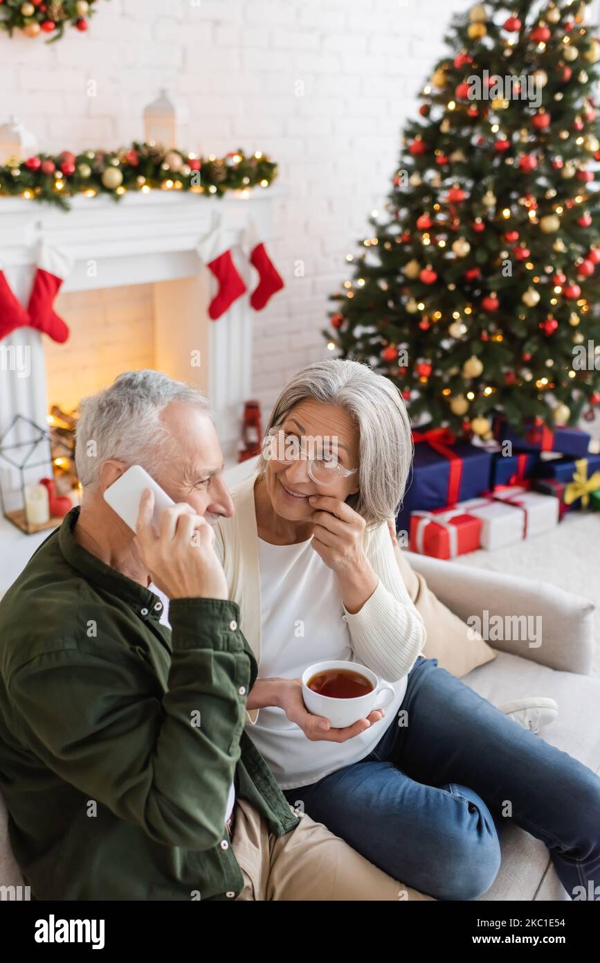 middle aged man talking on smartphone near curious wife with cup of tea ...