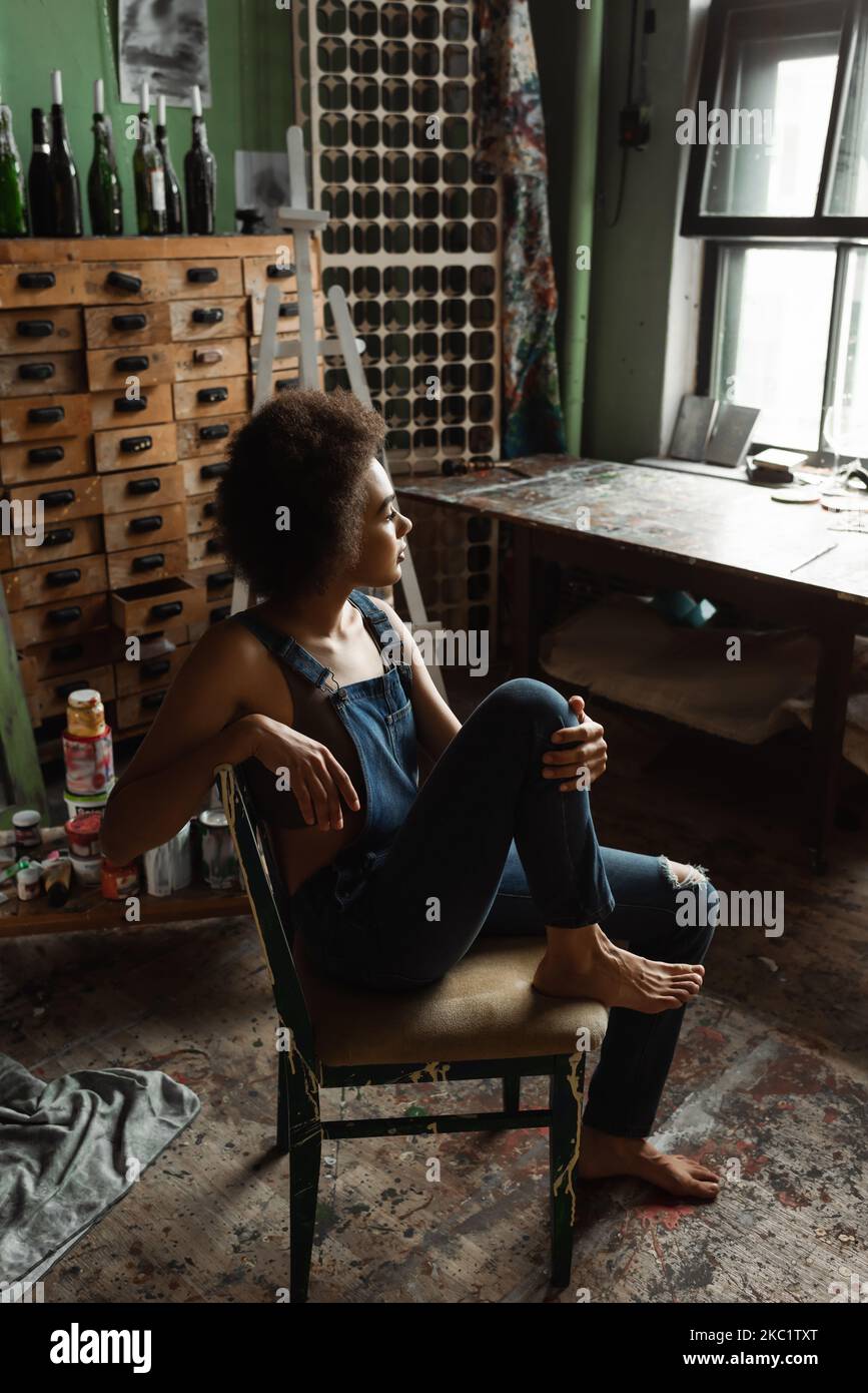 pretty and dreamy african american artist in overalls sitting barefoot ...