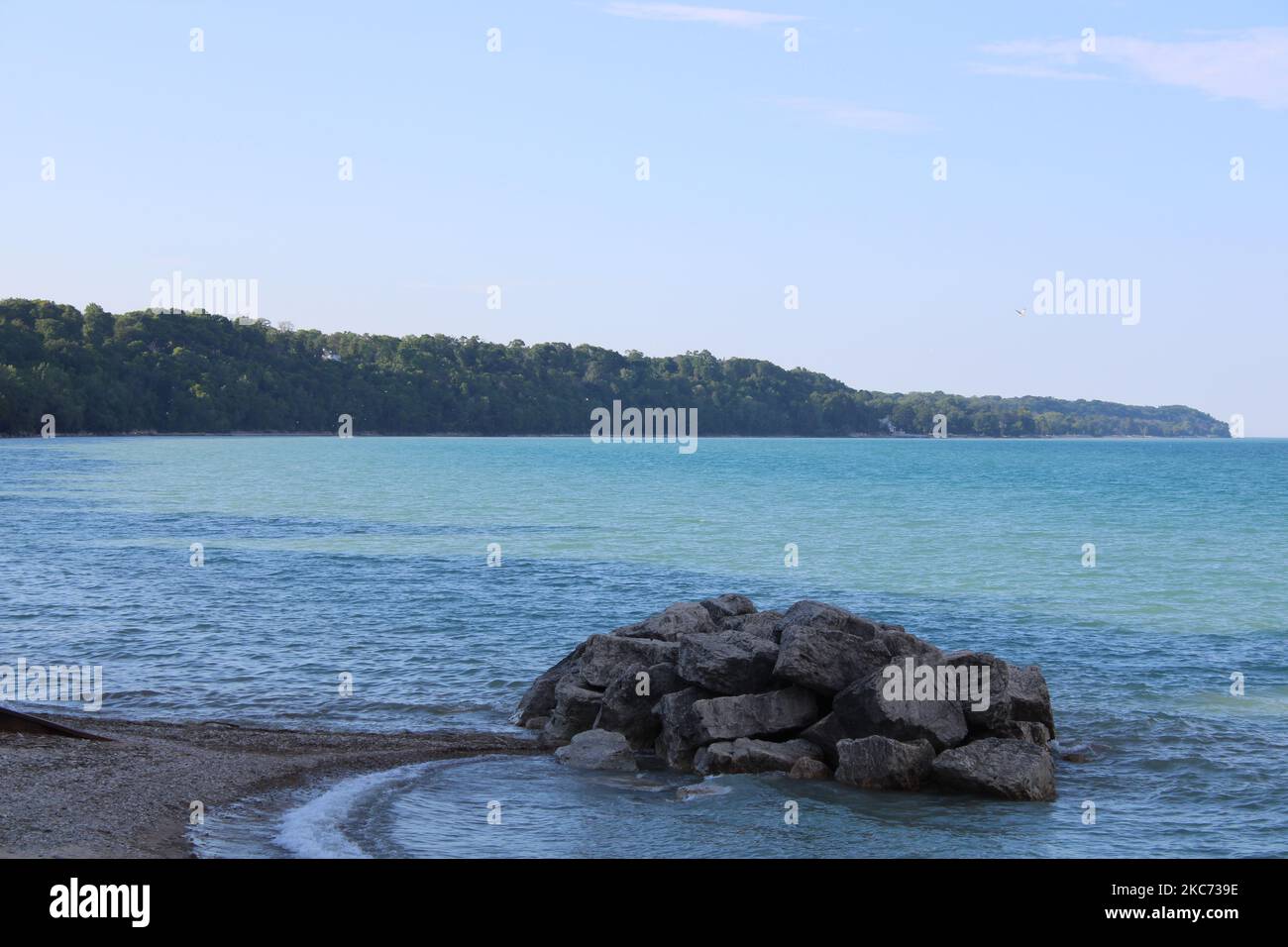 A stack of rocks on the lakeshore with clear blue water and green ...